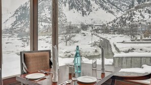Mesa posta com pratos, talheres, um copo e uma garrafa, com vista para um pico nevado pela janela.