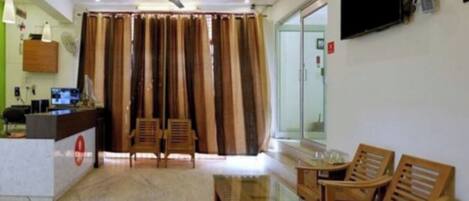Um hall de hotel com uma mesa de recepção, cadeiras de madeira, uma mesa com tampo de vidro, cortinas, um ventilador de teto e uma televisão fixada na parede.