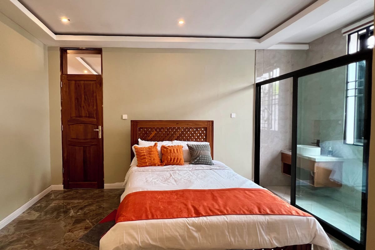 Quarto com cama, porta de madeira e porta de vidro deslizante que dá para um banheiro.