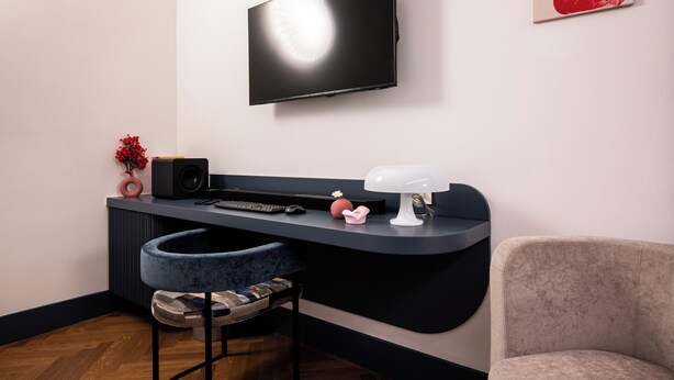 Quarto moderno com televisão de tela plana na parede, uma escrivaninha com cadeira, uma pequena mesa com um vaso e um alto-falante, e um sofá.