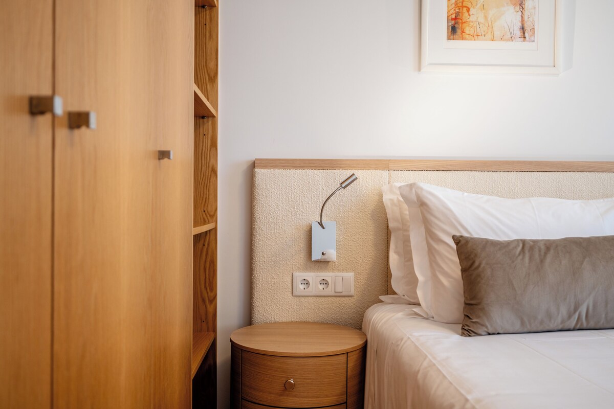 Quarto com guarda-roupa de madeira, mesa de cabeceira com abajur e cama com roupa de cama branca e travesseiro marrom.