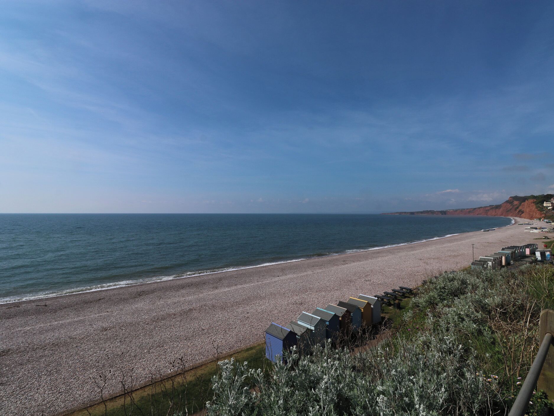 Uma praia costeira com areal de seixos, uma fileira de cabanas de praia e falésias ao fundo.