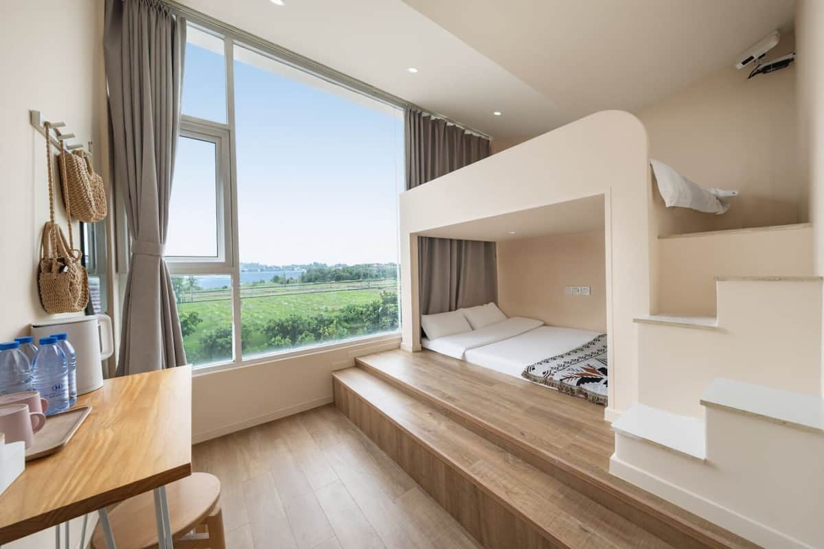 Quarto com beliche, escrivaninha, janela com vista e uma sacola pendurada.