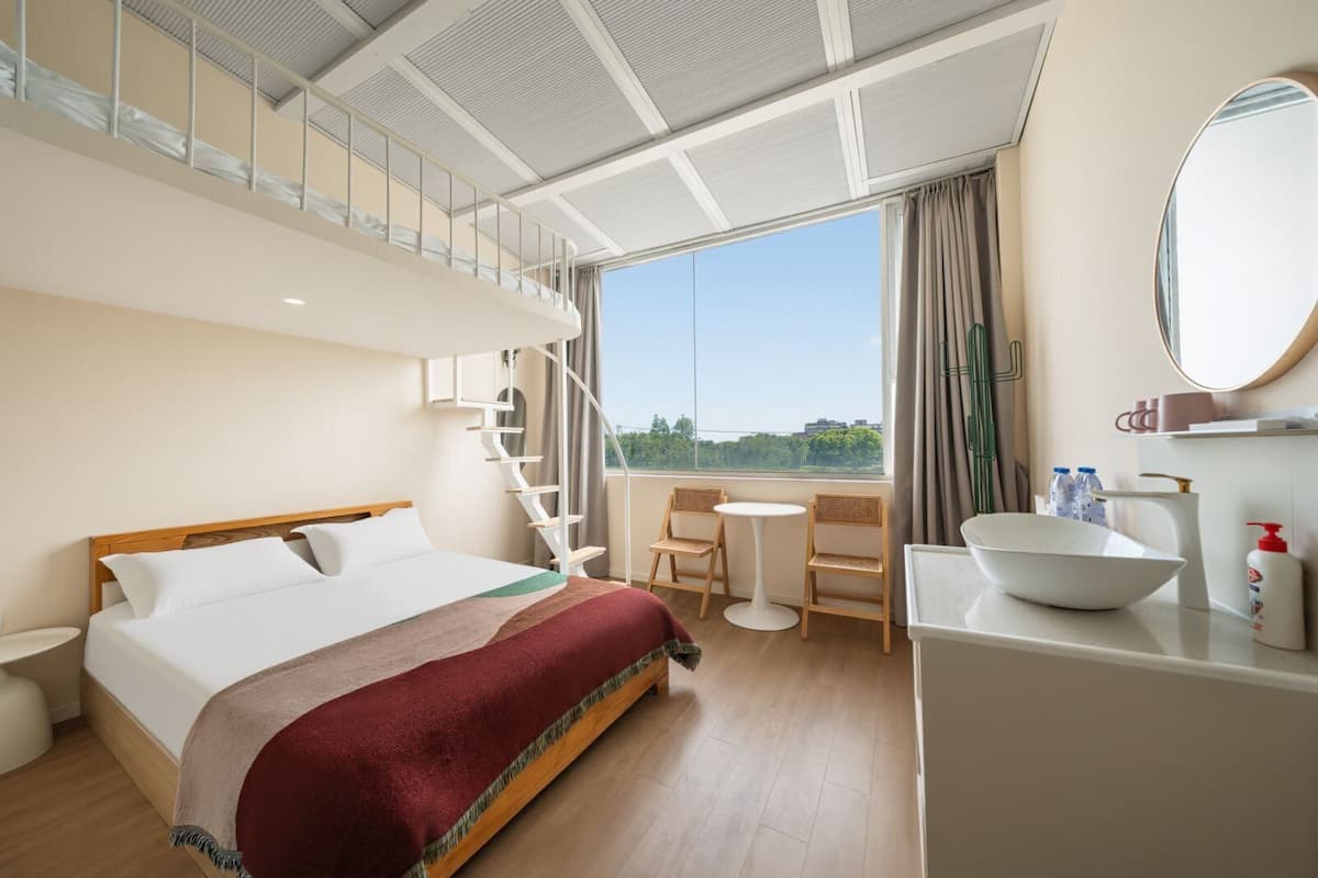 Quarto de hotel com cama, escrivaninha com pia, duas cadeiras, espelho redondo e vista para o verde lá fora.