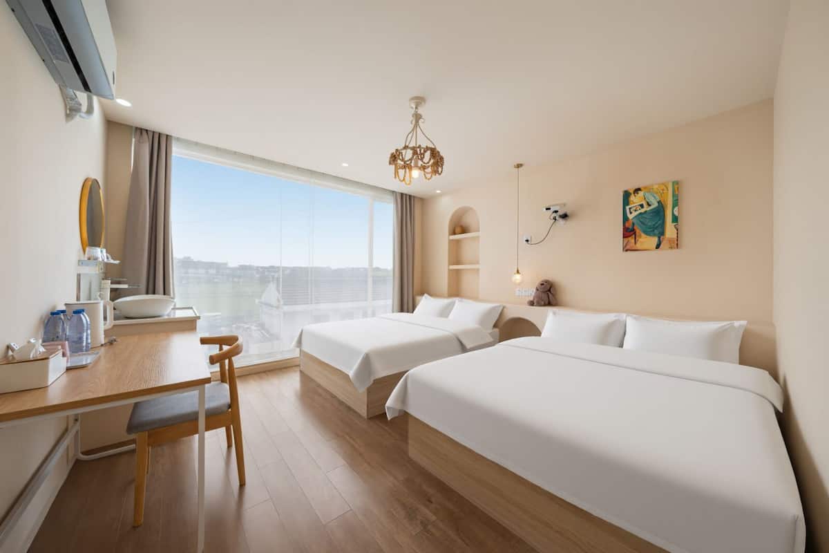 Quarto de hotel com escrivaninha, duas camas, um candelabro, um quadro e uma janela grande.