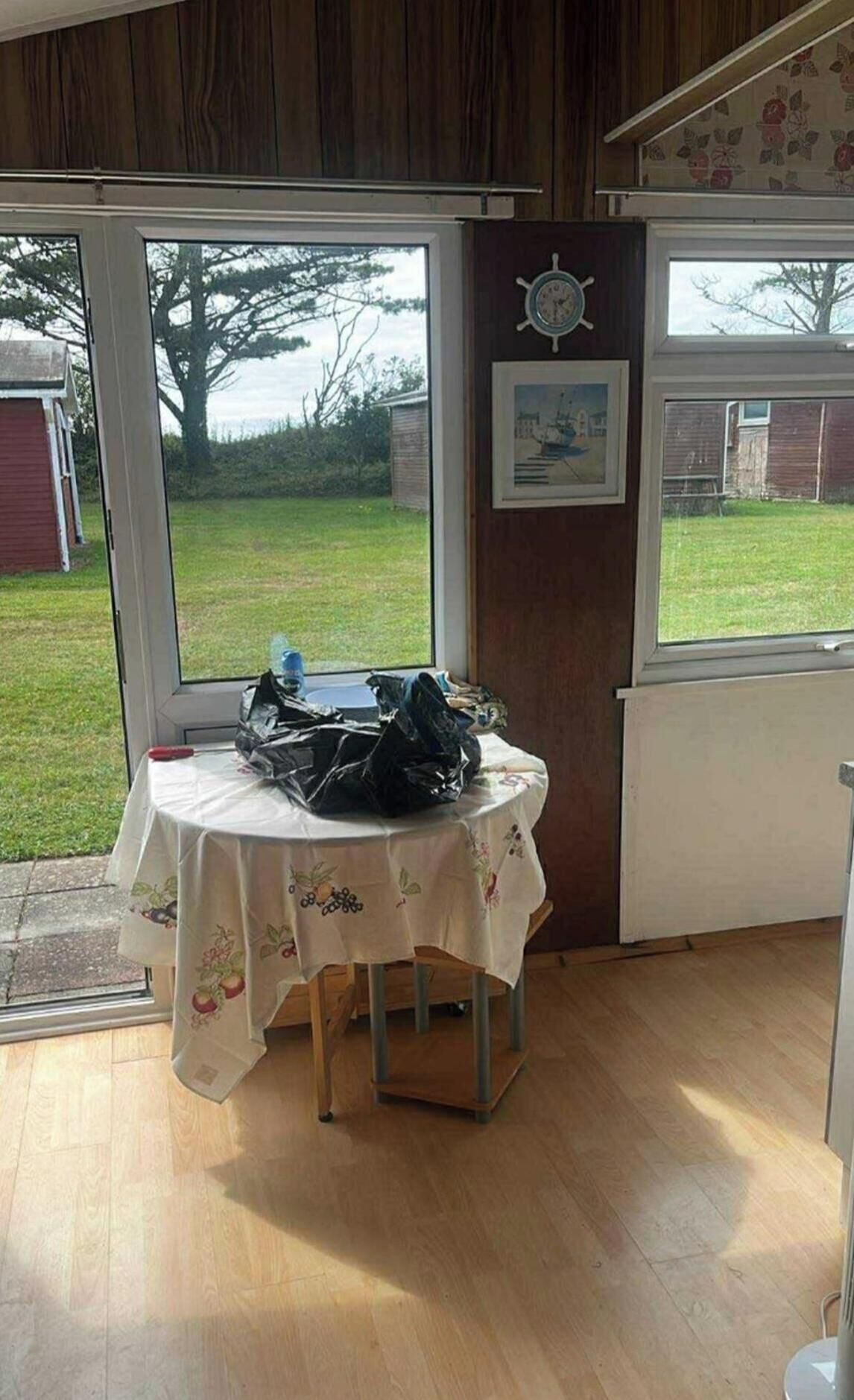 A room with a table covered by a tablecloth, a clock on the wall, and a window with a view of a grassy yard.