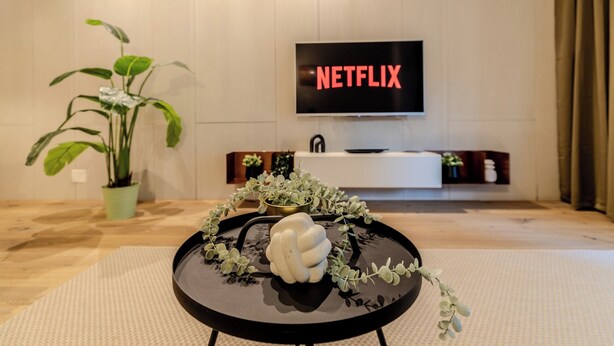 Uma sala de estar moderna com uma televisão de tela plana exibindo 'NETFLIX', uma mesa de centro redonda preta com um objeto decorativo branco e plantas, além de dois vasos com plantas.