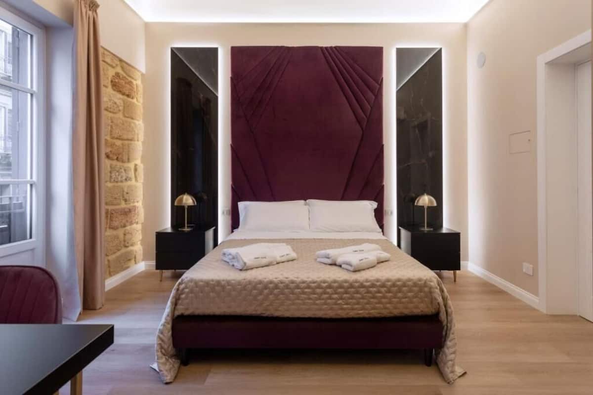Uma cama com cabeceira, duas mesas de cabeceira, uma janela e uma parede de pedra.