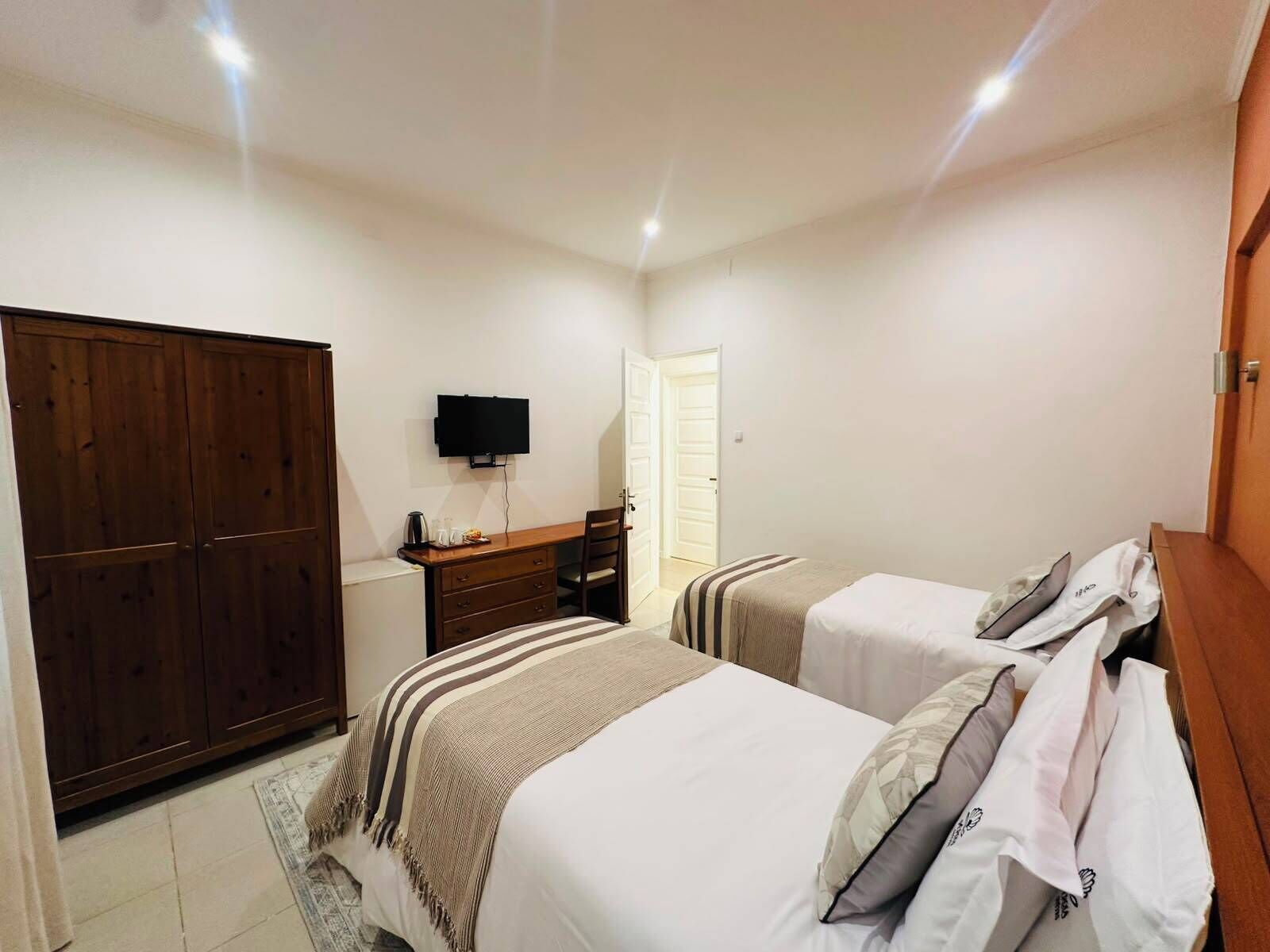 Quarto com duas camas, um guarda-roupa, uma escrivaninha com televisão e uma porta que dá para outro cômodo.