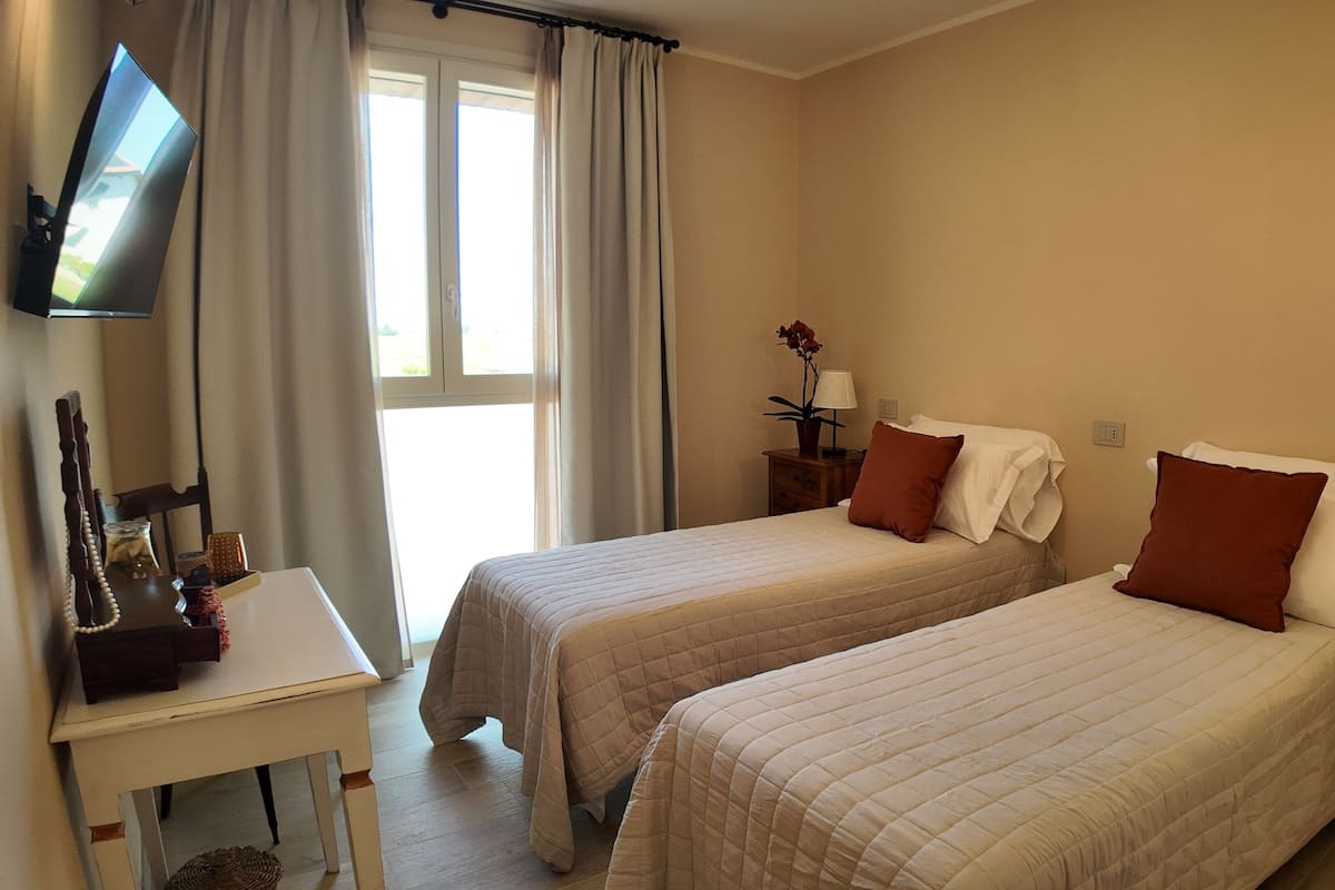Quarto de hotel com duas camas, uma escrivaninha, televisão e janela com cortinas.