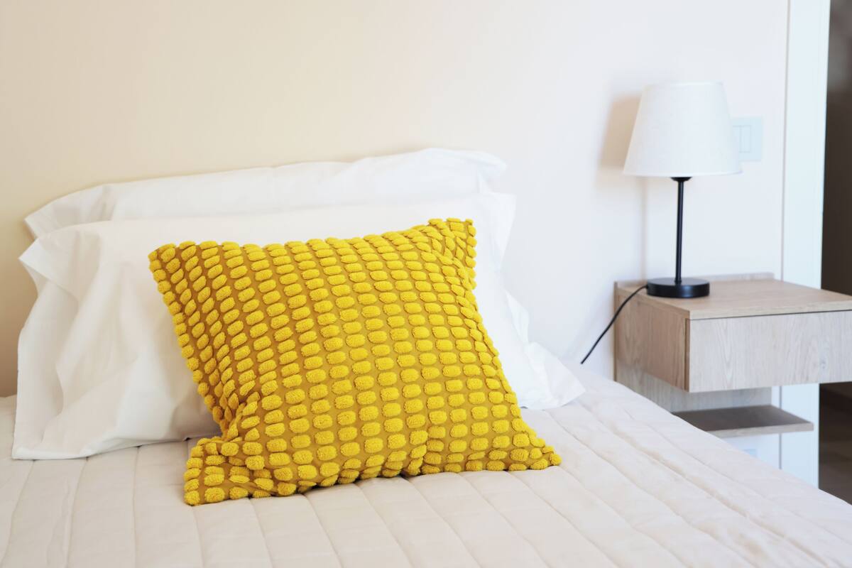 Cama com roupa de cama branca e almofada amarela texturizada, mesa de cabeceira com abajur e mesa de apoio de madeira.