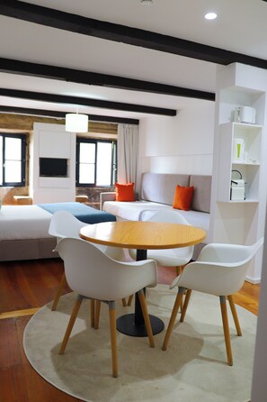 Uma sala de estar moderna com uma mesa de madeira redonda cercada por cadeiras brancas, um sofá com almofadas laranja e uma televisão fixada na parede.
