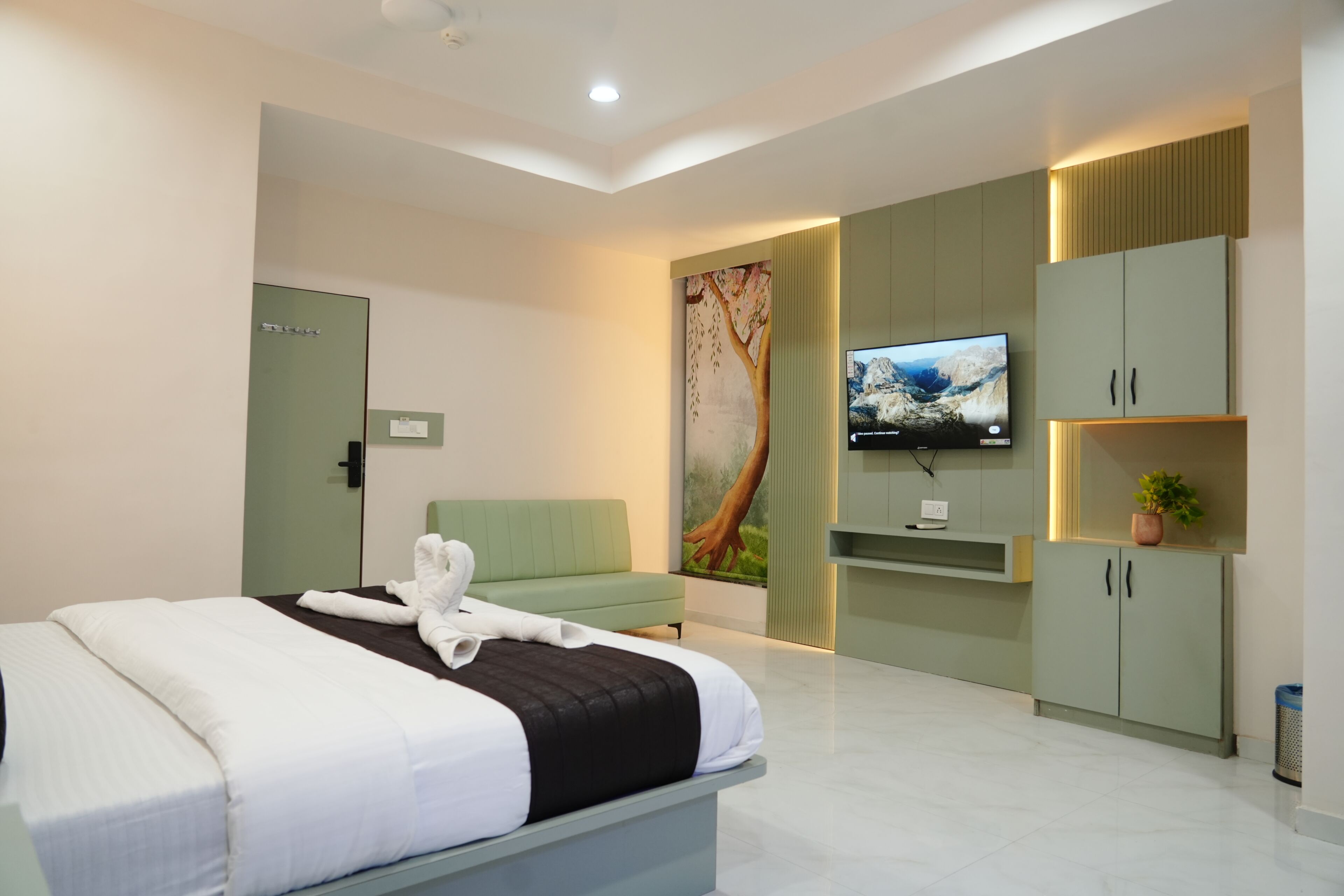 Quarto de hotel moderno com uma cama, televisão de tela plana, sofá verde, televisão fixada na parede e um armário.