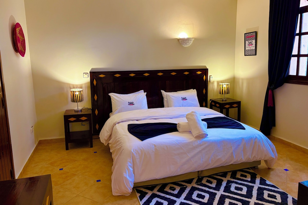 Quarto de hotel com cama, duas luminárias de cabeceira, arandela, tapete estampado e janela com cortinas.