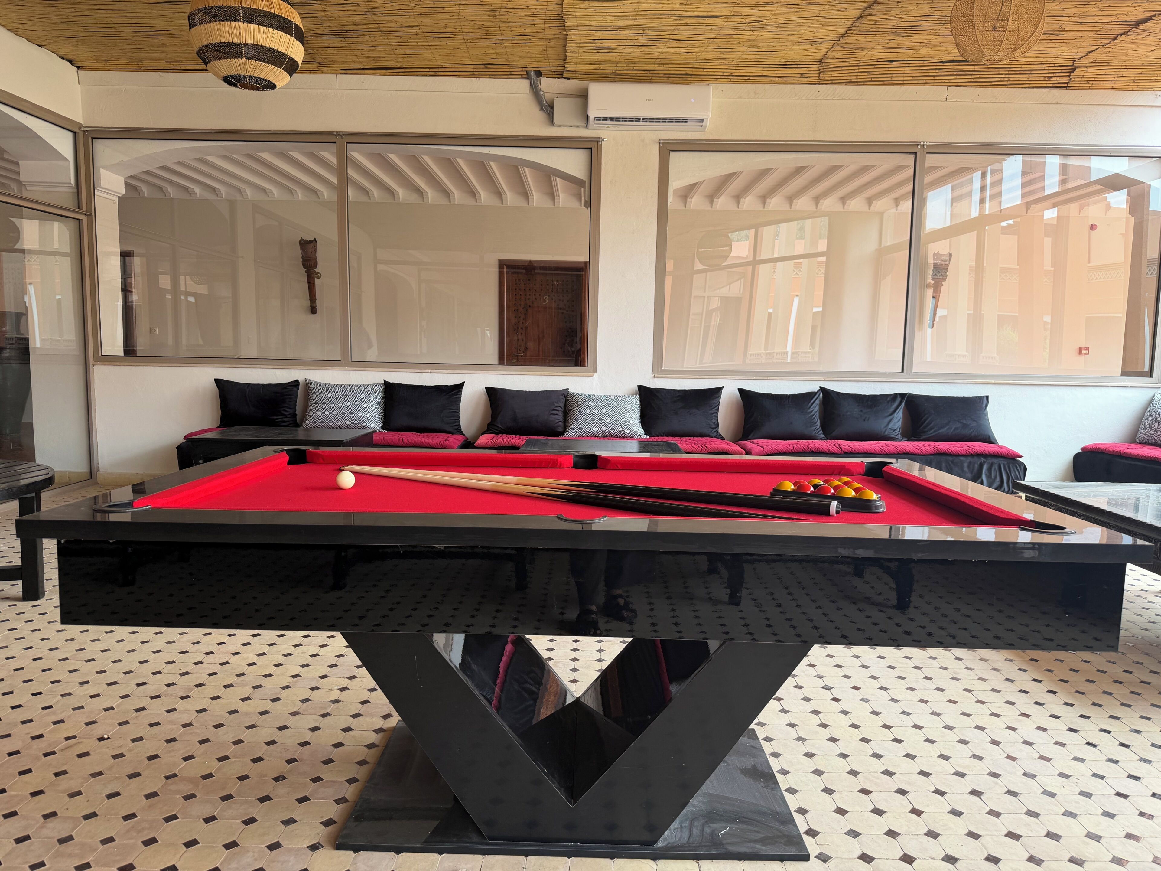 Uma mesa de bilhar com tecido vermelho, amortecedores pretos e um taco, num espaço com soalho estampado e grandes janelas.