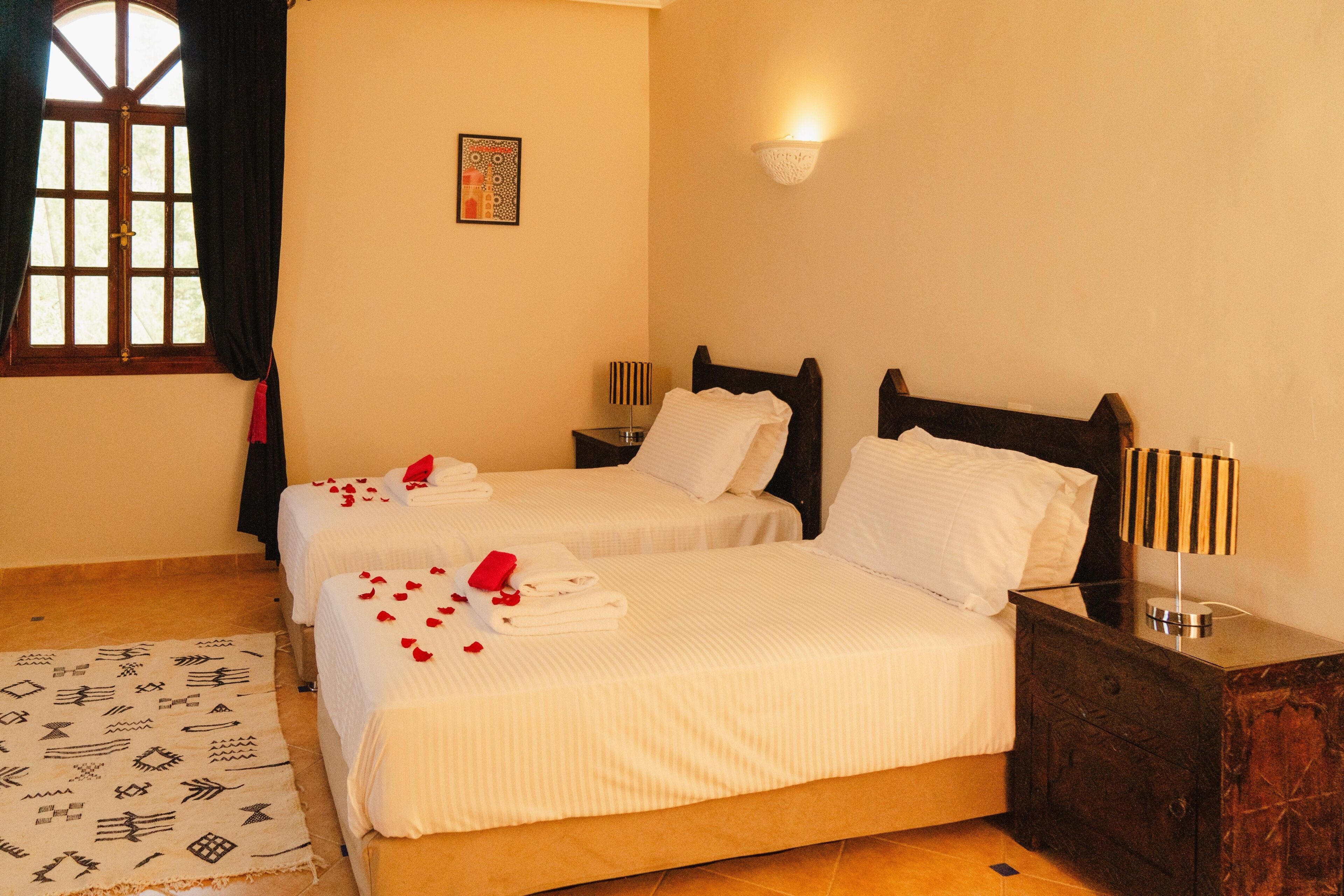 Quarto de hotel com duas camas, um tapete estampado, uma janela com cortinas, uma luminária na parede e uma mesa pequena com abajur.