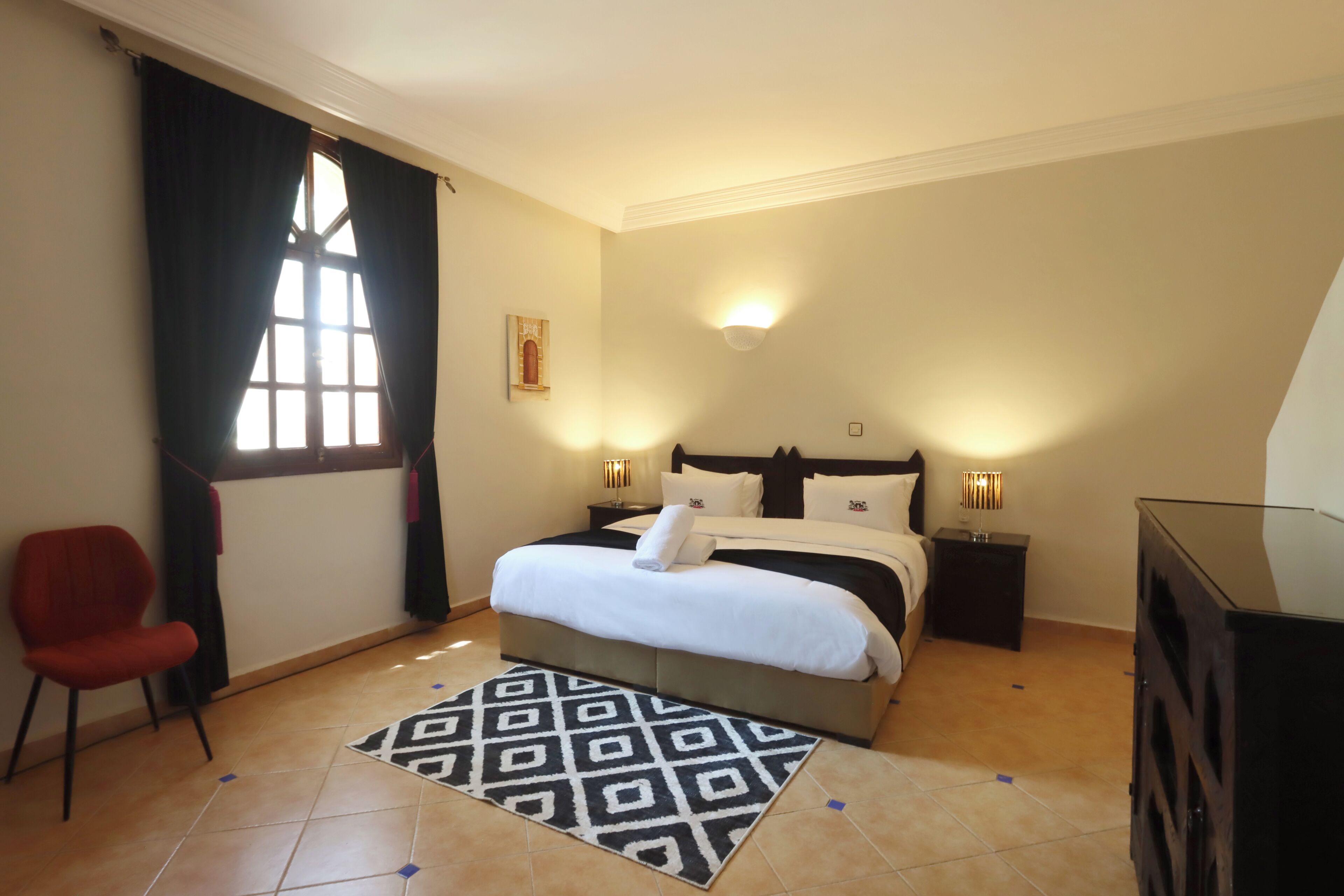 Quarto de hotel com cama, luminárias de cabeceira, uma cadeira vermelha, tapete estampado e um quadro na parede.