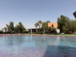 Piscina com superfície de água azul e límpida, rodeada por vegetação e um edifício com estilo arquitetônico tradicional.
