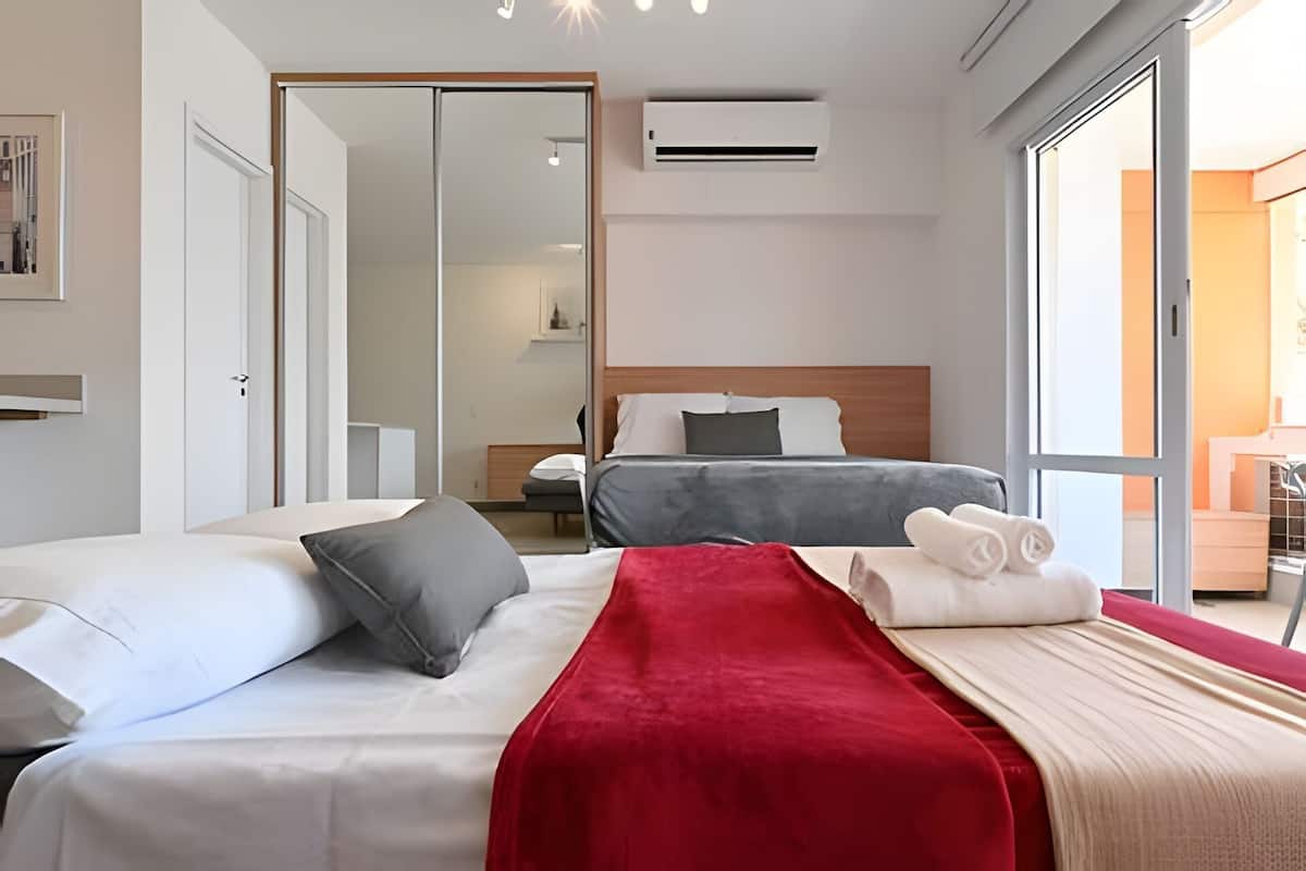 Quarto de hotel com duas camas, manta vermelha, toalhas brancas, cabeceira de madeira e ar condicionado.