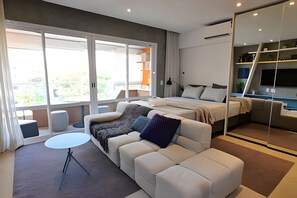 Quarto de hotel moderno com sofá, cama, mesinha e uma janela grande.