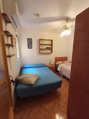 Quarto com uma cama de solteiro, mesa de cabeceira, ventoinha no teto e prateleira na parede.