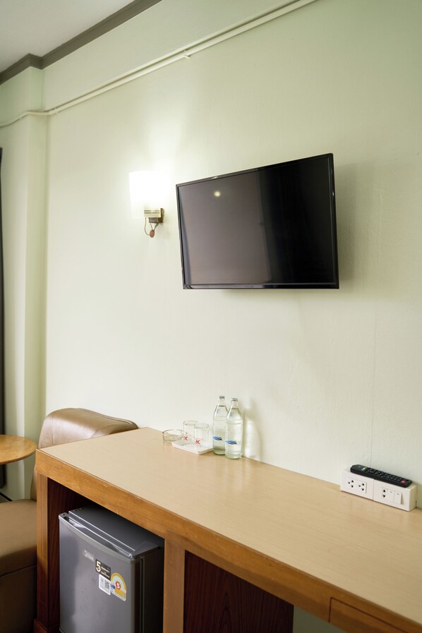 Quarto de hotel com televisão de tela plana na parede, uma escrivaninha de madeira com geladeira e um controle remoto sobre a escrivaninha.