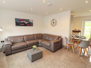 Uma sala de estar com sofá seccional, mesa de centro, área de jantar com mesa e cadeiras, relógio de parede e uma pintura de carro esportivo.
