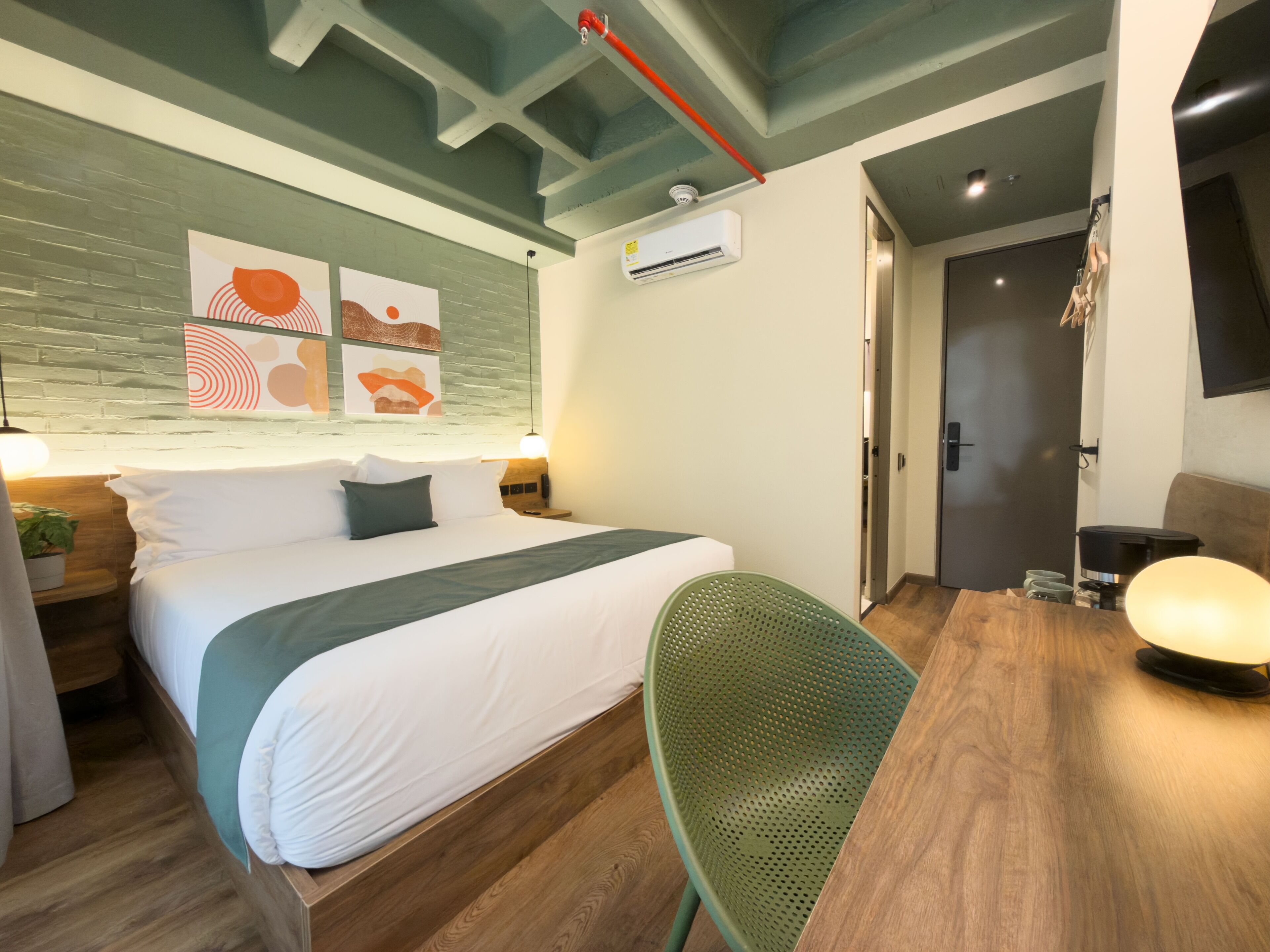 Quarto de hotel com uma cama, uma cadeira verde, uma mesa de madeira e obras de arte na parede.