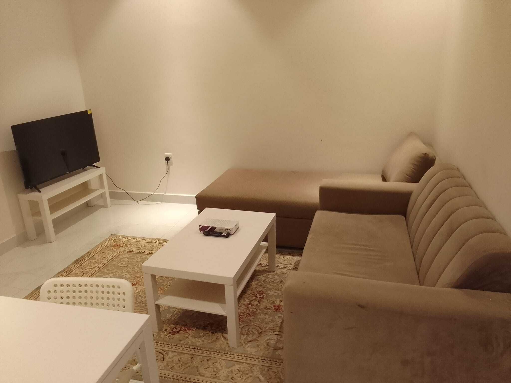 Uma sala de estar com uma televisão de tela plana em um suporte, um sofá bege, uma mesa de centro branca e um tapete estampado.