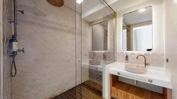Casa de banho com chuveiro envolto em vidro, lavatório branco com cuba e espelho sobre o lavatório.