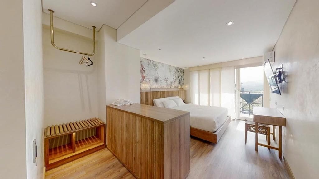 Quarto de hotel moderno com escrivaninha de madeira, cama com roupa de cama branca, janela grande e TV fixada na parede.