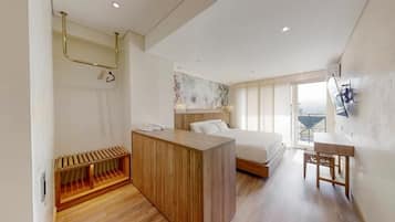 Quarto de hotel moderno com escrivaninha de madeira, cama com roupa de cama branca, janela grande e TV fixada na parede.