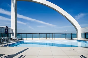 Piscina no terraço com vista clara para o mar, rodeada por arquitetura moderna e um guarda-corpo de vidro.