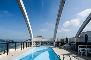 Piscina no terraço com vista desobstruída do oceano e um edifício moderno ao fundo.