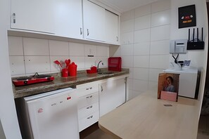Uma cozinha com armários brancos, bancada de granito, chaleira elétrica vermelha e uma geladeira pequena.