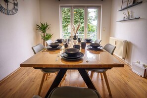 Uma área de refeições com uma mesa de madeira posta para quatro pessoas, rodeada por cadeiras, com uma janela que oferece vista para o verde lá fora.