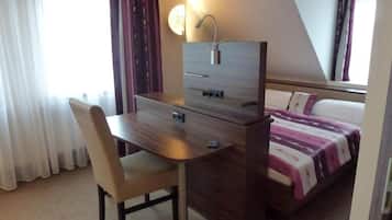 Quarto de hotel com cama, escrivaninha com cadeira, mesa de cabeceira e janela com cortinas.
