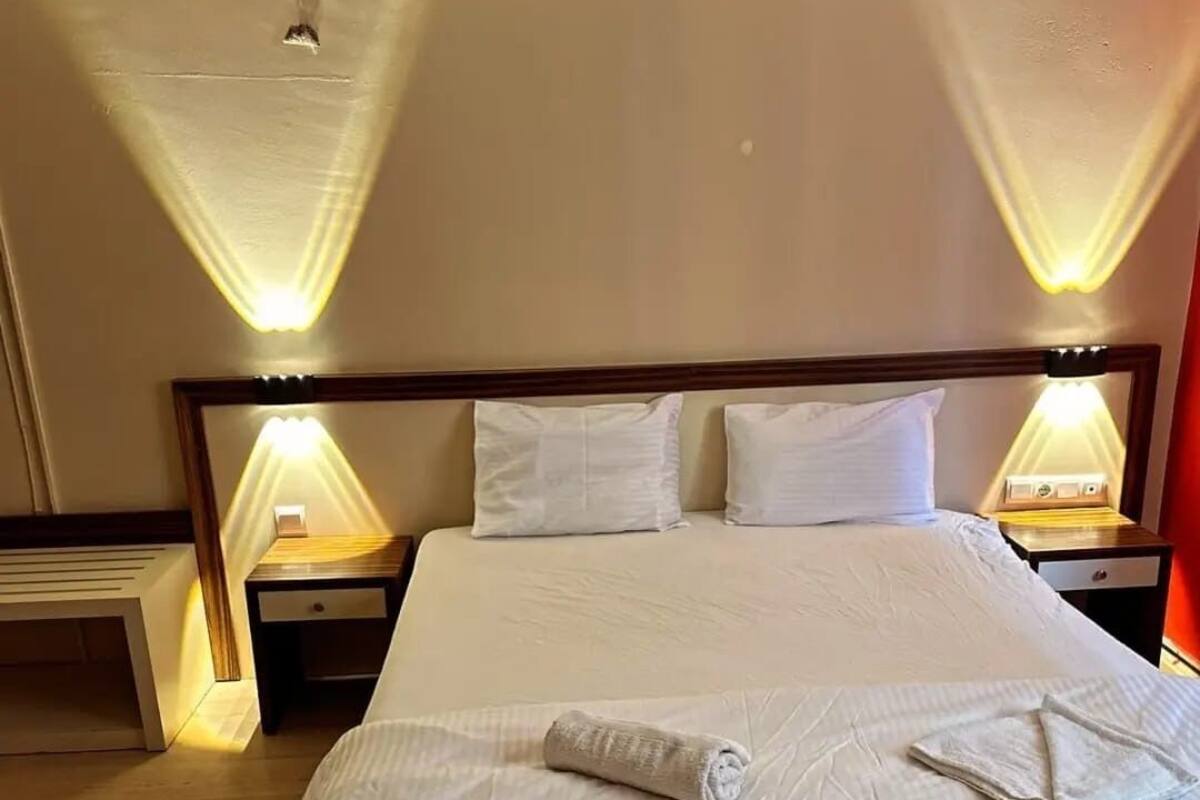 Uma cama bem arrumada com lençóis e almofadas brancas, duas mesas de cabeceira com abajures e um banco de madeira.