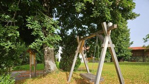 Um conjunto de baloiços de madeira num parque, com árvores e uma pequena construção de madeira ao fundo.