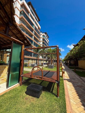 Área externa cercada com banco, uma mesa pequena e um caminho que leva a um edifício com várias varandas.