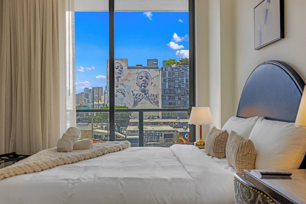 Quarto de hotel com uma cama grande, vista para a cidade, um quadro na parede e uma mesa de cabeceira com abajur.