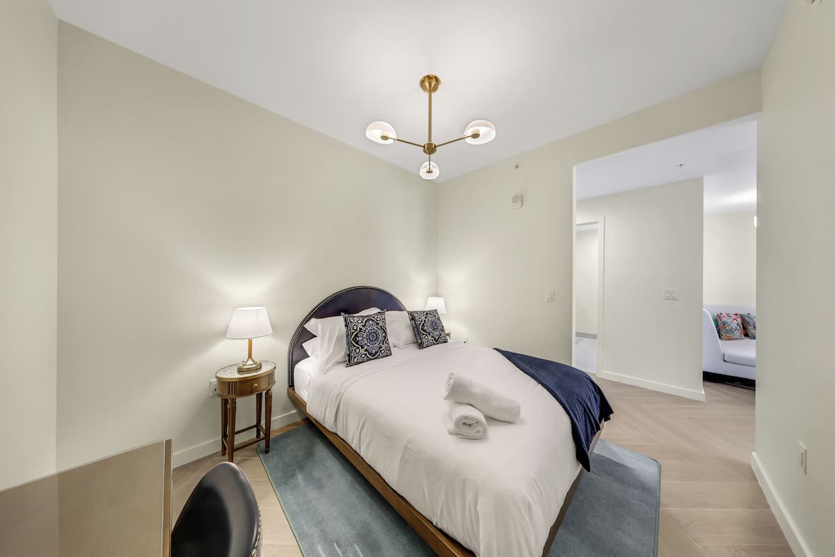 Um quarto com cama, mesas de cabeceira, abajur, ventilador de teto e uma porta que dá para outro cômodo.
