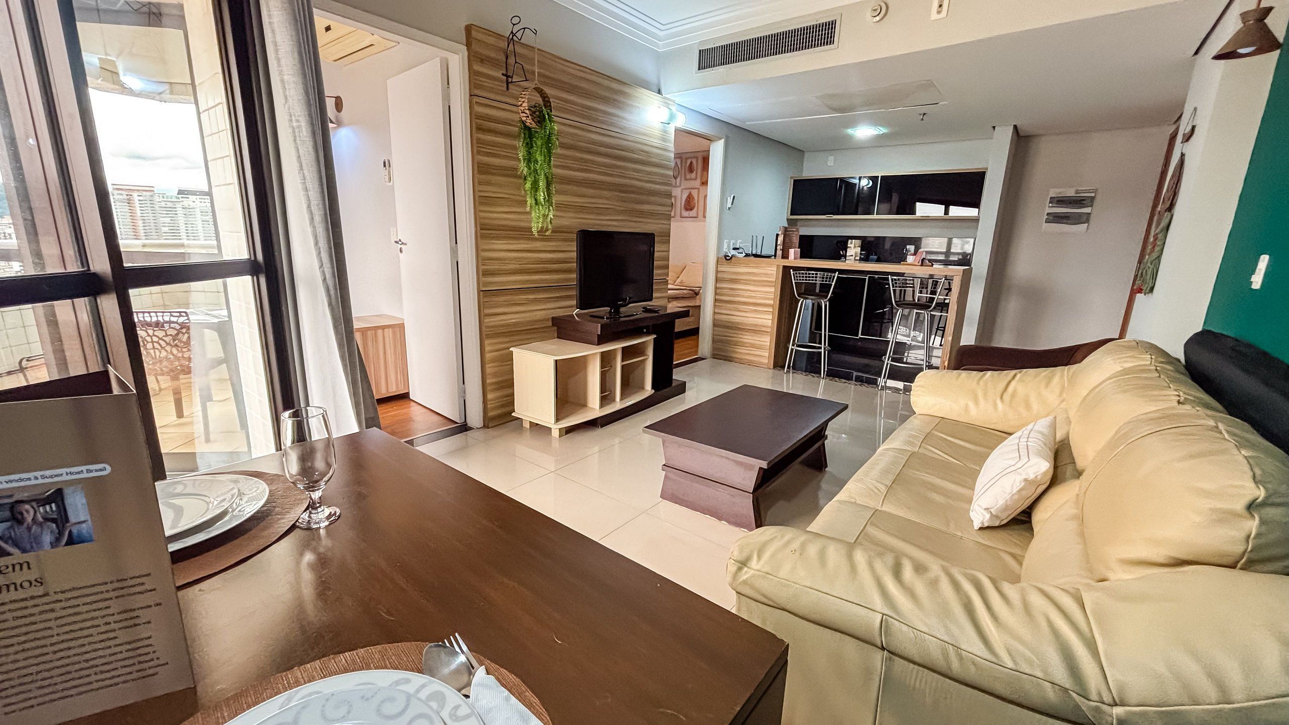 Uma sala de estar moderna com um sofá, mesa de centro e uma TV. É visível uma área de cozinha ao fundo.