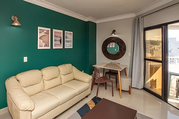 Sala de estar com um sofá bege, uma mesinha e uma cadeira. Há três quadros na parede, um espelho redondo e uma janela com vista para prédios.