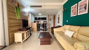 Sala de estar com um sofá bege, uma TV em um suporte e uma cozinha ao fundo.