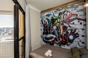 Quarto com um mural de super-heróis na parede, uma cama e uma janela com vista para a paisagem urbana.