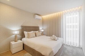 Quarto com cama, mesas de cabeceira, abajures, ar condicionado e cortinas leves.