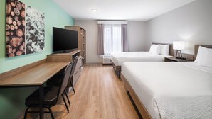 Quarto de hotel com secretária de madeira, televisão de tela plana, duas camas, janela com cortinas e obras de arte na parede.