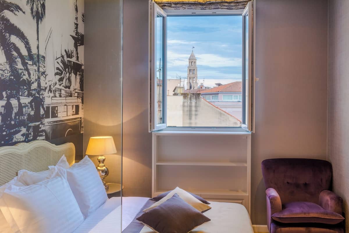 Quarto de hotel com cama, cadeira, janela e vista para um edifício, além de uma luminária.