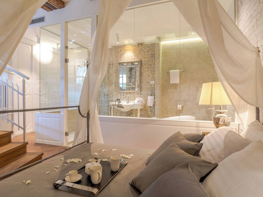 Quarto com cama, bandeja com objetos, janela e banheiro visível pela porta de vidro.